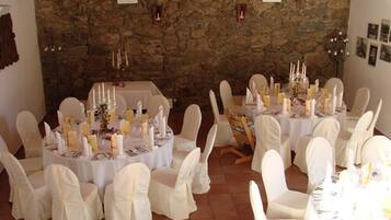 Salão de banquetes com mesas redondas preparadas para um evento formal, cadeiras brancas e um fundo de parede de pedra.