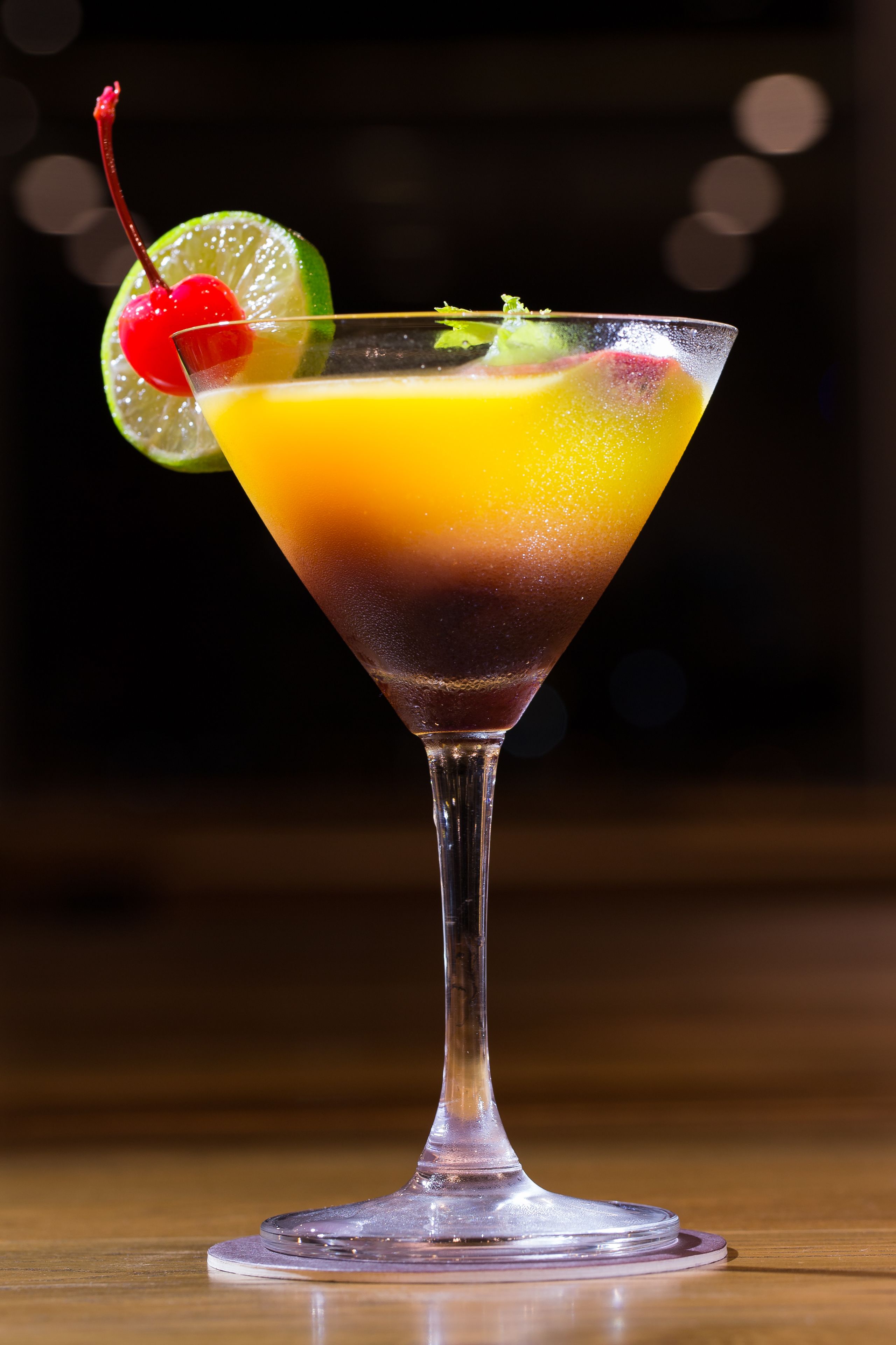 Um copo de martini com uma bebida em camadas, decorado com uma cereja e uma fatia de limão, sobre uma superfície de madeira.