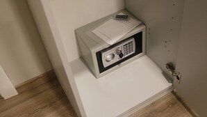 Um cofre pequeno, branco e com estrutura de metal, com um teclado e um controle remoto em cima, colocado sobre uma prateleira branca encostada a uma parede.