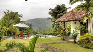 Área de piscina com mesa e cadeiras, rodeada por vegetação exuberante e ao fundo, uma montanha.
