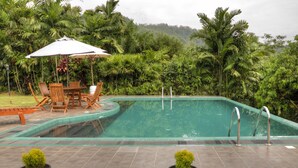 Piscina retangular com borda de vidro, rodeada por um deck ladrilhado. Há uma mesa e cadeiras de madeira com um guarda-sol branco, e duas plantas em vasos em primeiro plano.