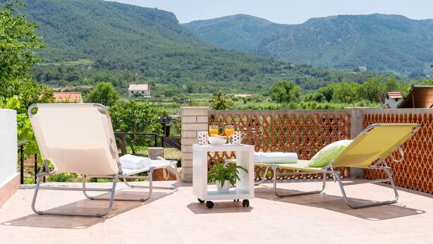 Um terraço com duas poltronas de descanso, uma pequena mesa com uma planta e um carrinho branco com uma tigela e bebida de laranja.