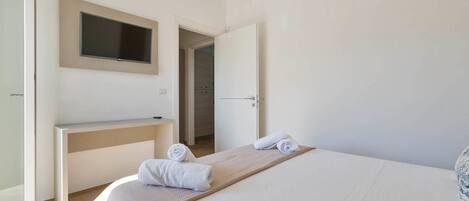 Quarto de hotel com cama, televisão fixada na parede, escrivaninha e banheiro visível por uma porta aberta.