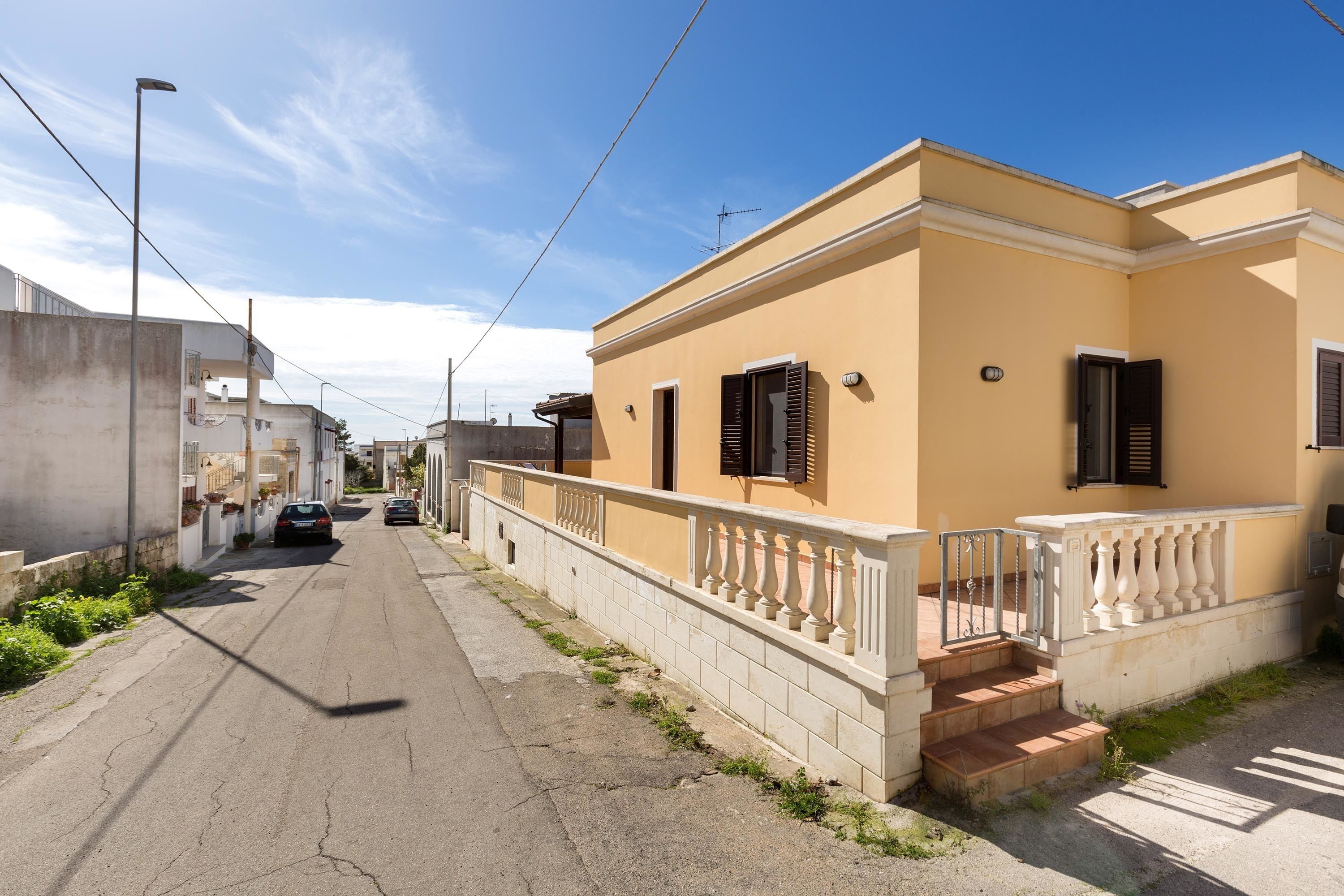 Uma rua estreita com um edifício amarelo à direita, apresentando uma varanda com balaustrada branca e um pequeno lance de degraus que levam à entrada.