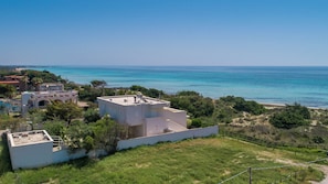 Um edifício costeiro com telhado plano, rodeado por vegetação e uma vista clara do mar.