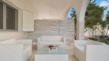 Uma área de estar moderna ao ar livre, com móveis de vime branco e uma mesa de centro de vidro, com um muro de pedra e vegetação ao fundo.