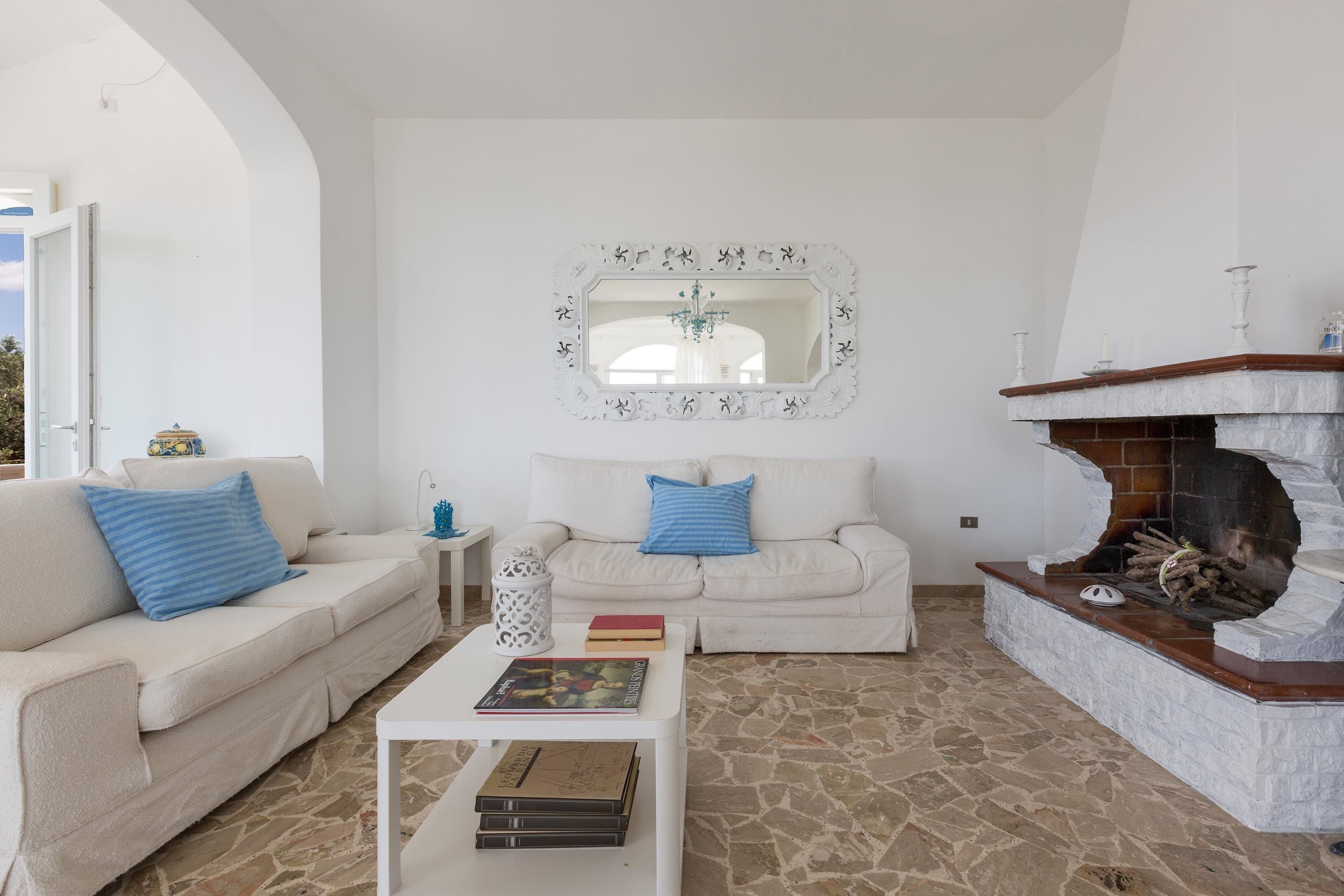 Sala de estar com dois sofás brancos, uma mesa de centro com livros, lareira e um espelho decorativo na parede.