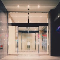 Entrada de um edifício moderno com porta de vidro, piso de azulejos e teto com iluminação embutida.