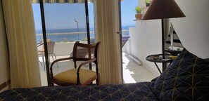 Varanda com vista para o mar, uma cadeira, uma mesa pequena com abajur e uma cama com colcha estampada.