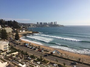 Paisagem costeira com praia, estrada com carros estacionados e um conjunto distante de edifícios altos.