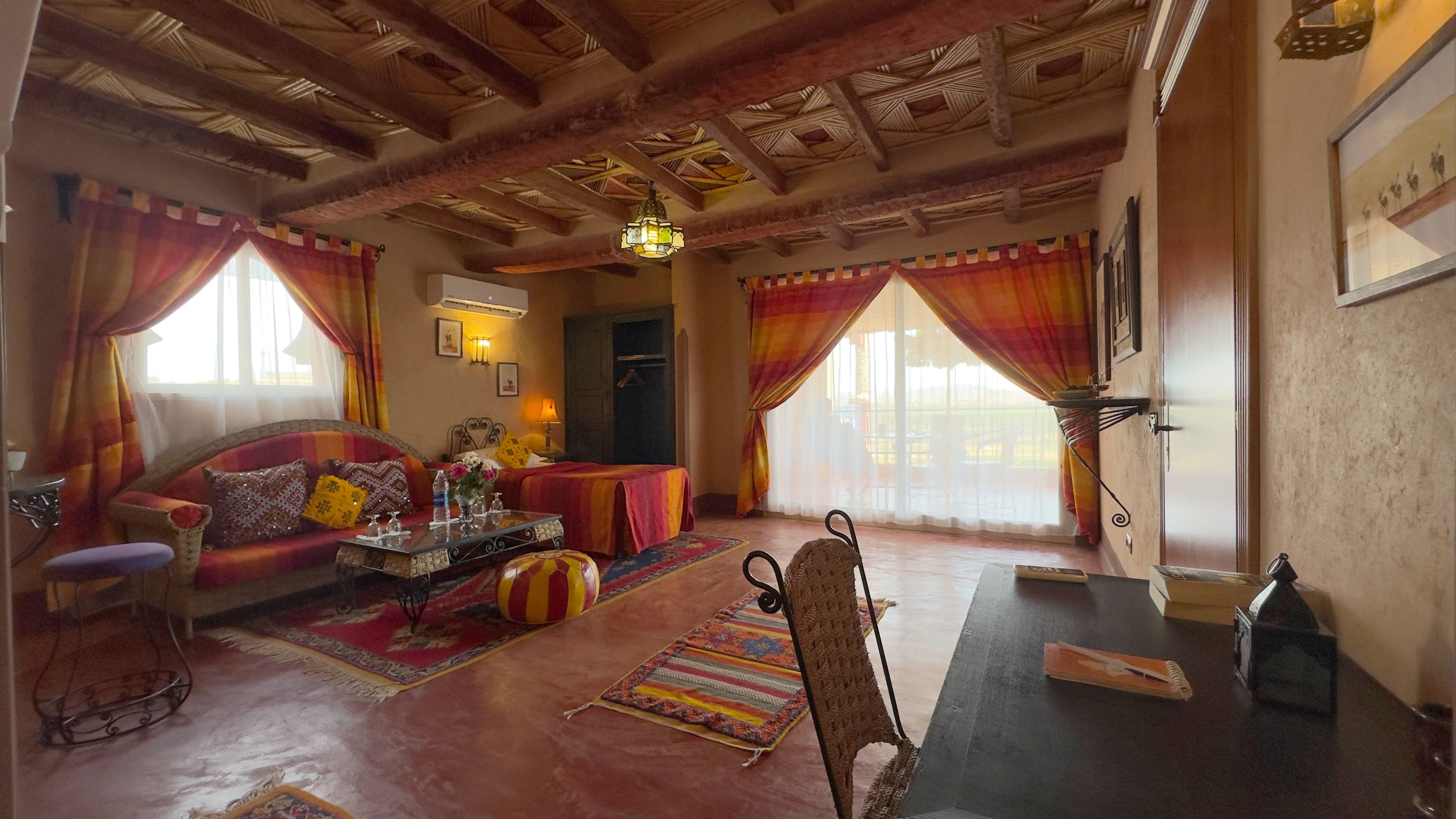 Uma sala de estar aconchegante com um sofá, uma mesa de centro e uma poltrona. Há uma janela com cortinas, um ventilador de teto e uma luminária na parede.