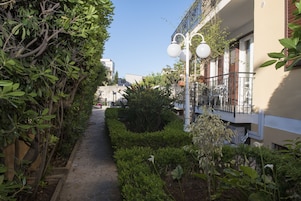Um caminho estreito ladeado por vegetação exuberante e um edifício com varandas e luminárias externas.