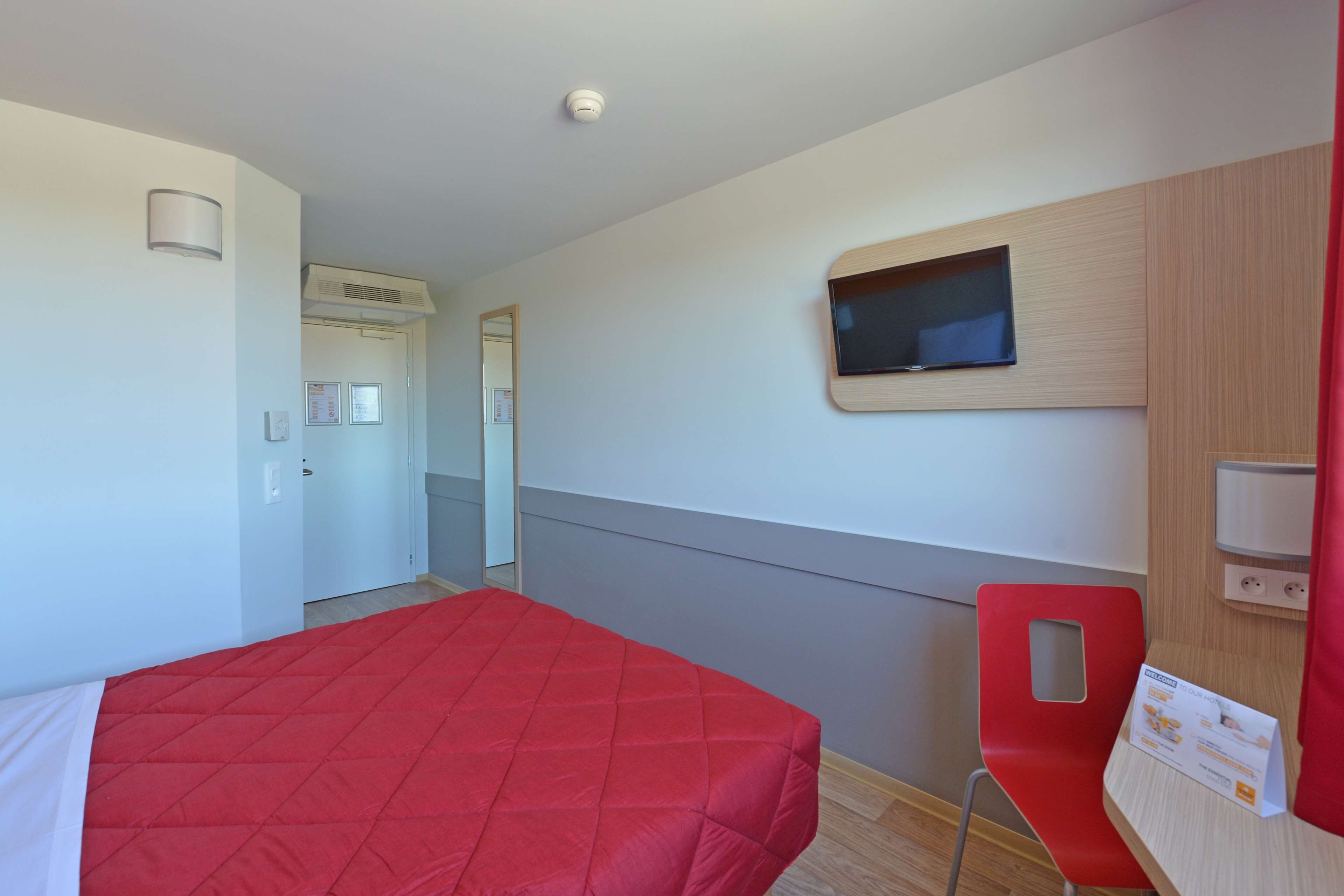 Quarto de hotel com cama vermelha, cadeira vermelha, televisão montada e uma pequena janela.