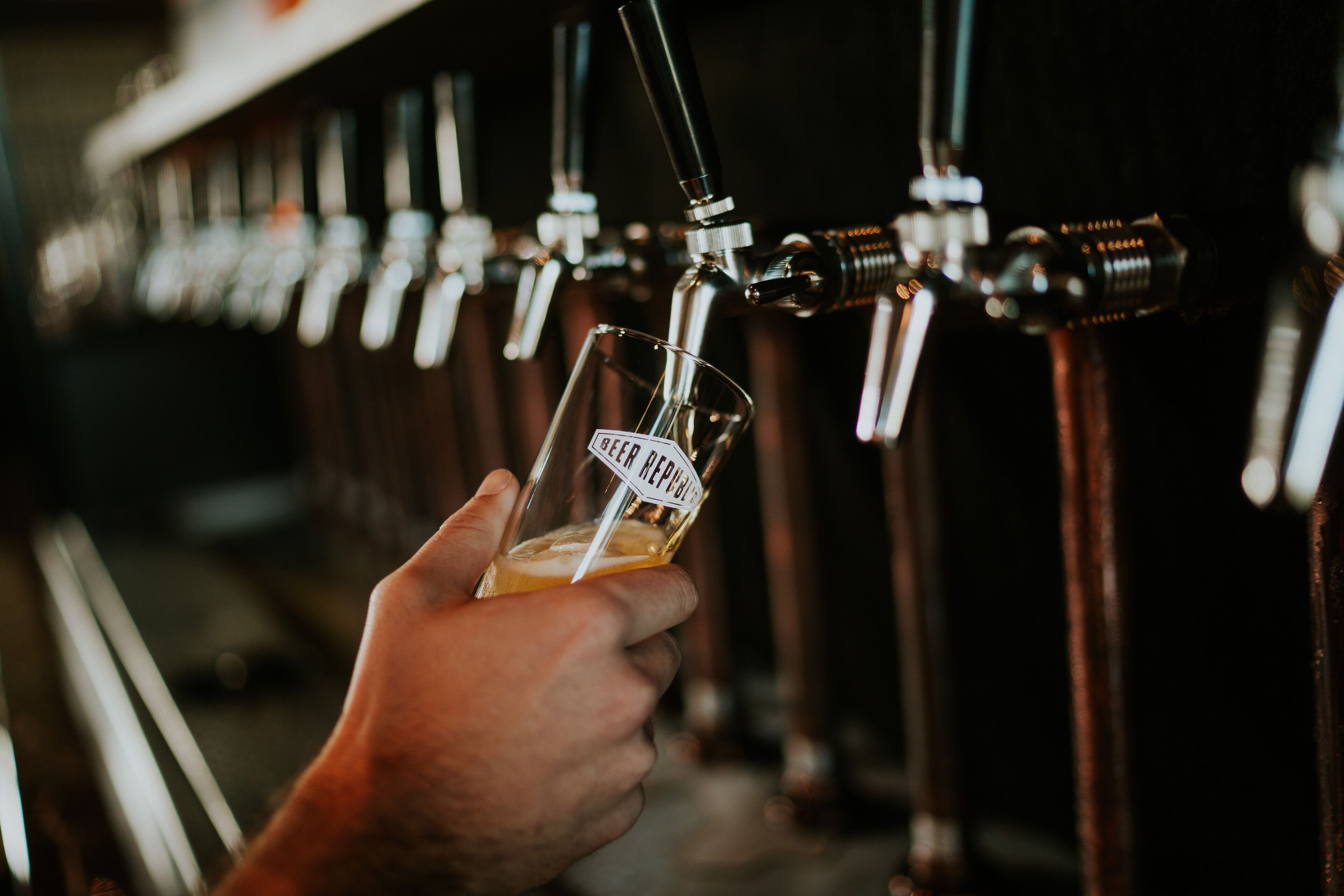 Uma mão segurando um copo de cerveja com uma etiqueta que diz 'BEER REPUBLIC' em um bar.