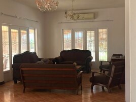 Uma sala de estar espaçosa com um sofá grande, outro menor e uma mesa de centro de madeira. Há duas janelas com detalhes em vidro decorativo e um aparelho de ar condicionado fixado na parede.