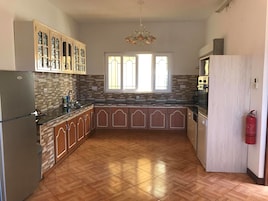 Cozinha equipada com frigorífico, fogão e pia. Há uma janela com três painéis e um candeeiro.