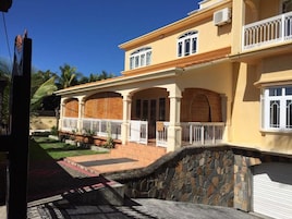 Uma casa de dois andares com um muro de suporte de pedra, um alpendre coberto com grades brancas e um céu azul claro.