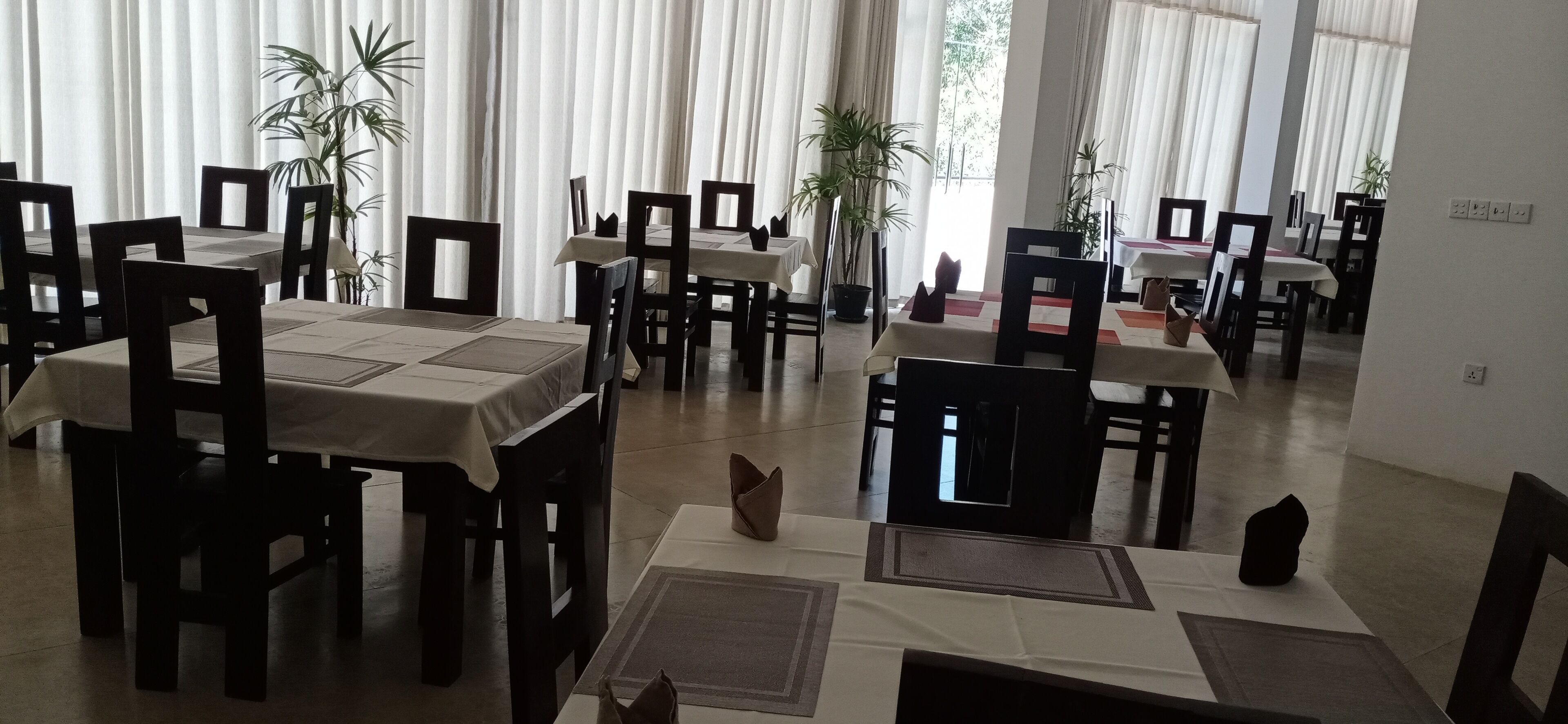 Uma sala de jantar com mesas e cadeiras, janelas amplas com cortinas e plantas em vasos.