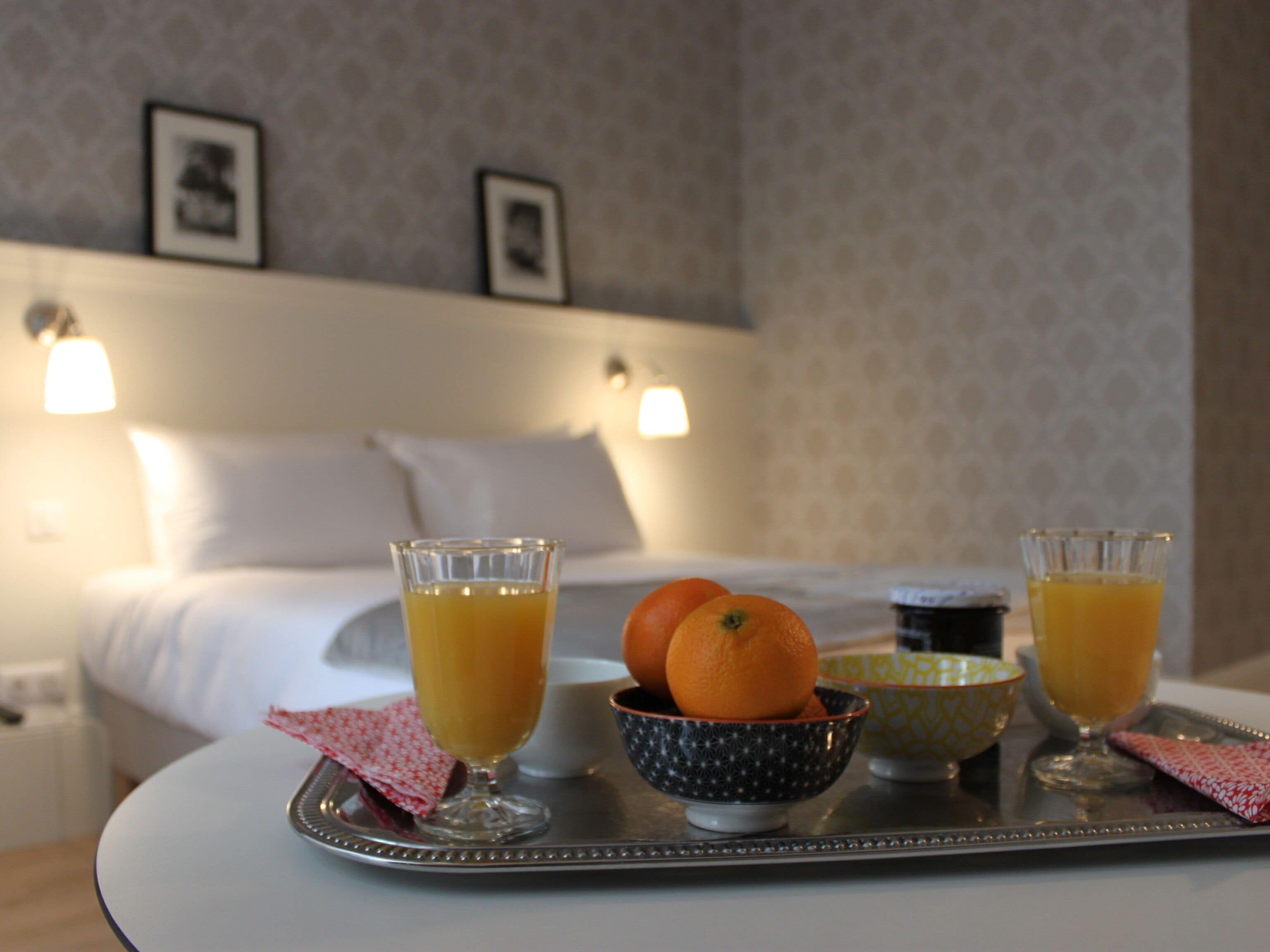 Bandeja de café da manhã com copos de suco de laranja, uma tigela de laranjas e uma bandeja decorativa em quarto de hotel, com cama e luminárias na parede.