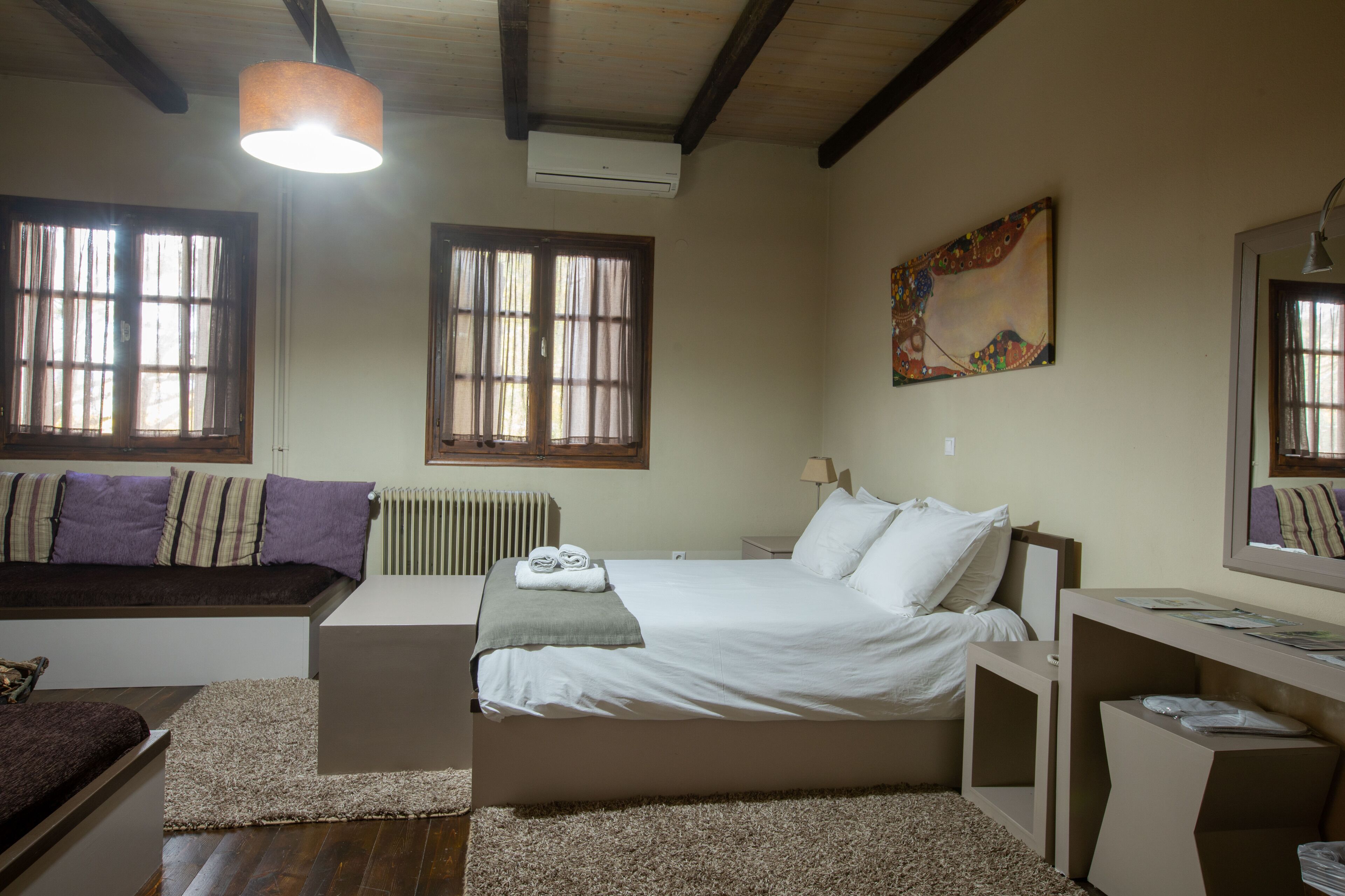 Quarto com cama, mesa de cabeceira e escrivaninha com cadeira. Há janelas com cortinas, um radiador e um quadro na parede.