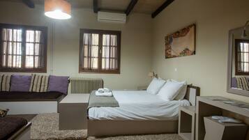 Quarto com cama, mesa de cabeceira e escrivaninha com cadeira. Há janelas com cortinas, um radiador e um quadro na parede.