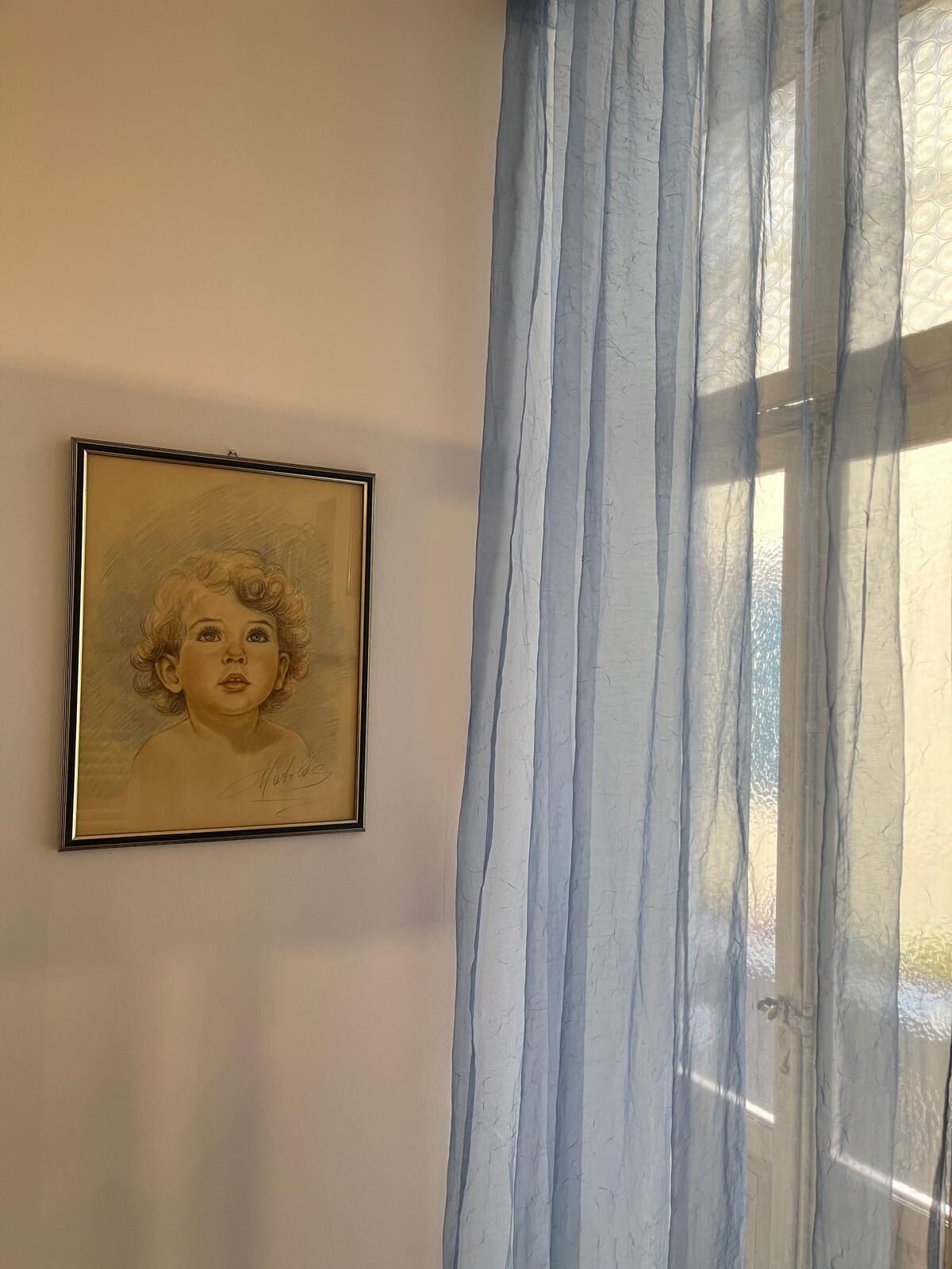 Uma obra de arte emoldurada numa parede, uma janela com cortinas translúcidas e um vislumbre do interior de um quarto.