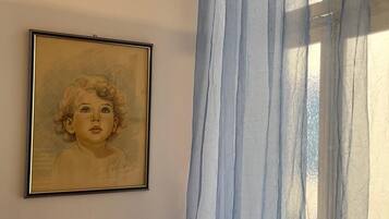 Uma obra de arte emoldurada numa parede, uma janela com cortinas translúcidas e um vislumbre do interior de um quarto.