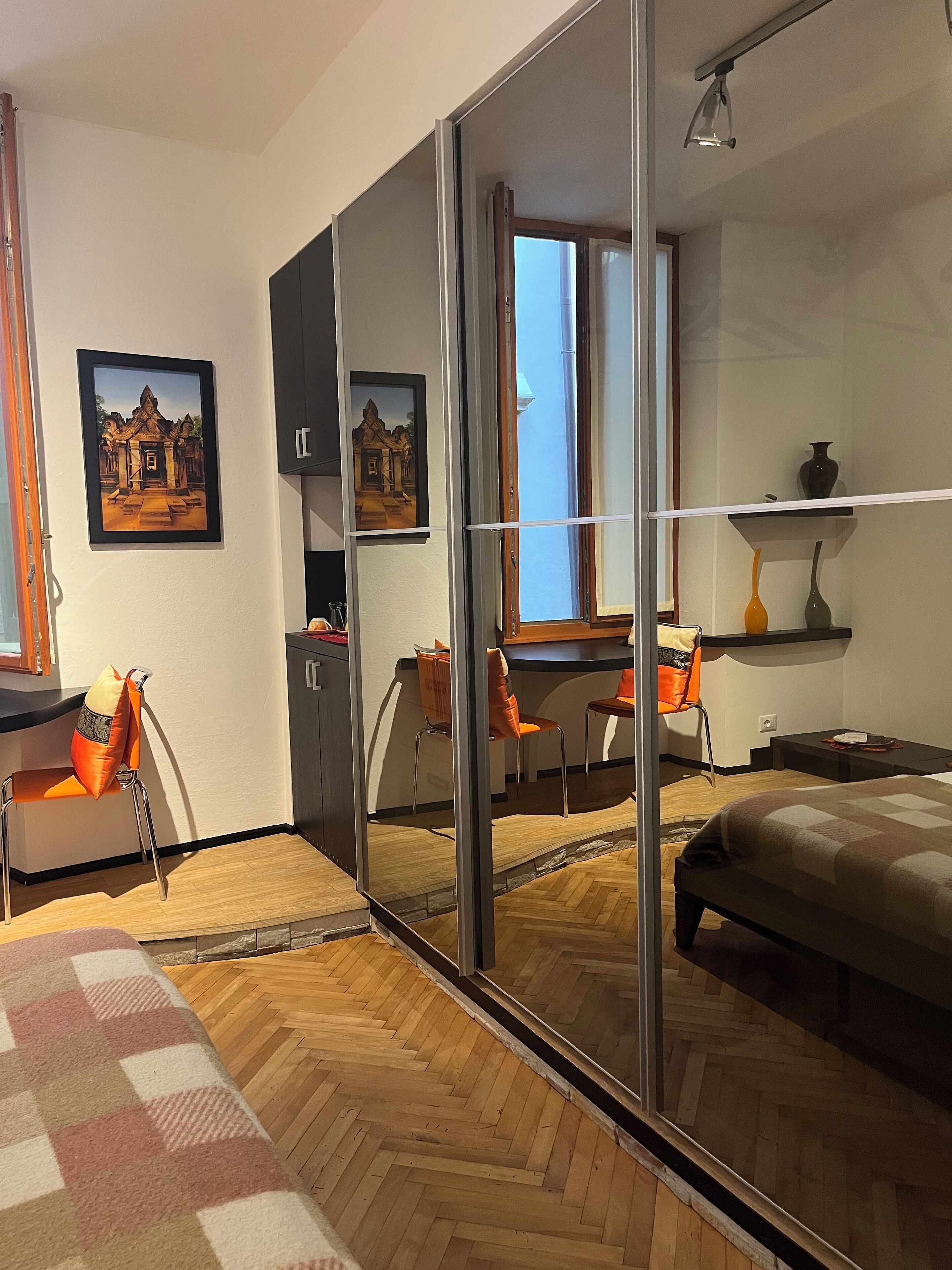 Quarto de hotel moderno com um espelho grande, uma cama, uma escrivaninha e uma cadeira. Há duas molduras com quadros na parede e uma janela com cortinas.