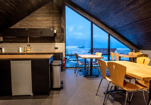 Cozinha moderna com uma grande janela para o oceano, mesas e cadeiras de madeira e uma pequena geladeira.