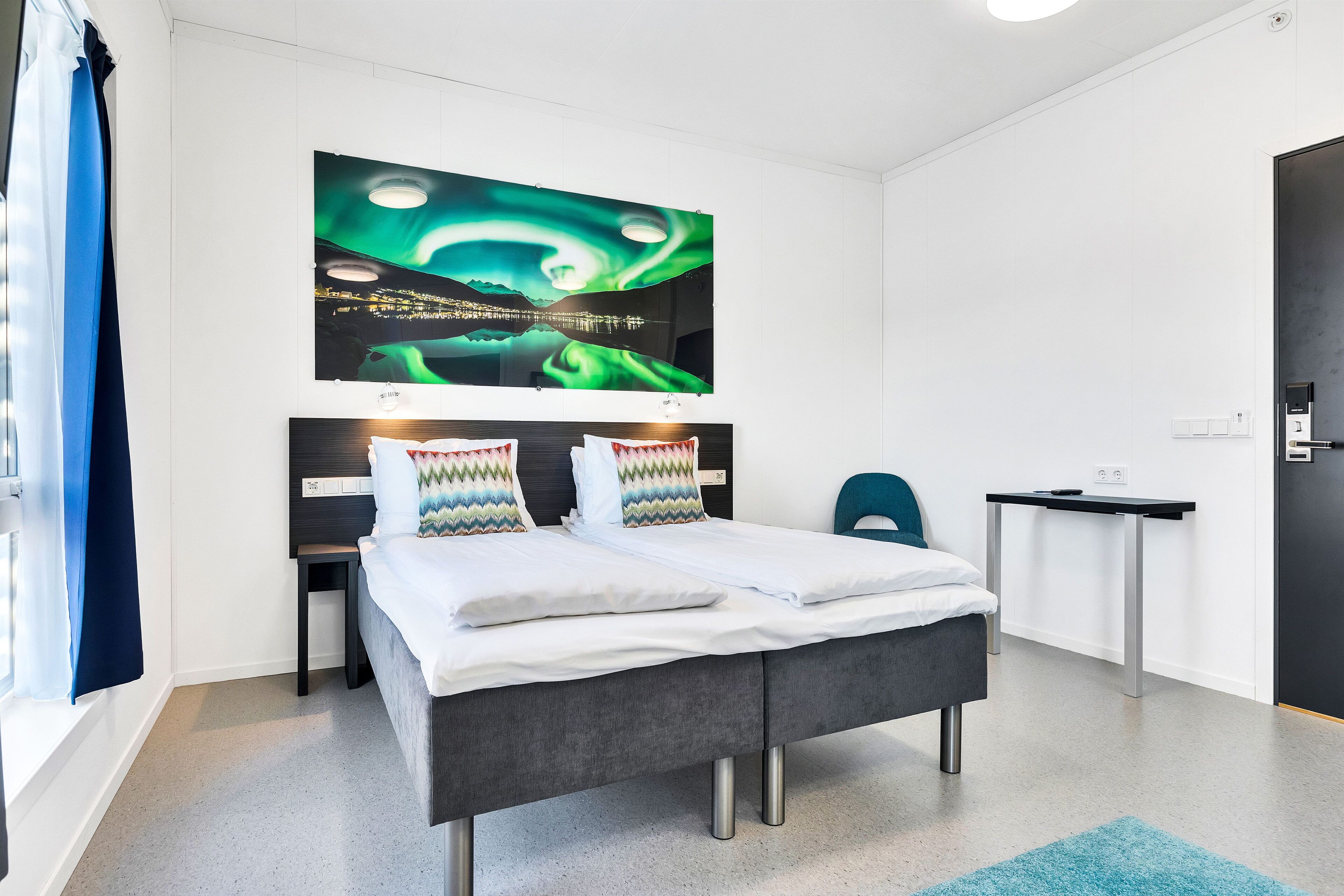 Quarto de hotel moderno com uma cama, uma escrivaninha e uma grande obra de arte da aurora boreal na parede.