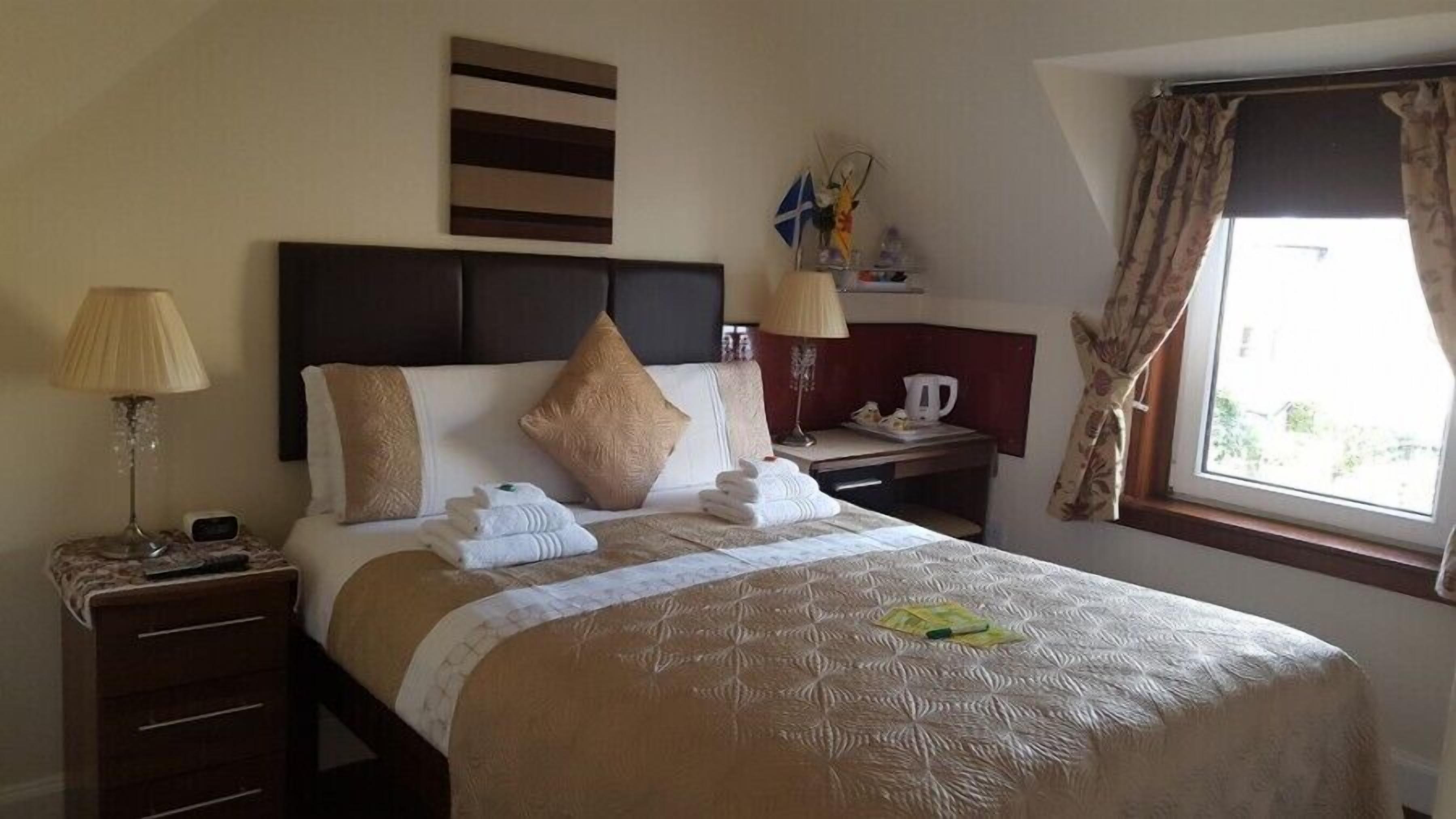 Quarto de hotel com cama, mesas de cabeceira, abajur, cafeteira e janela com cortinas.