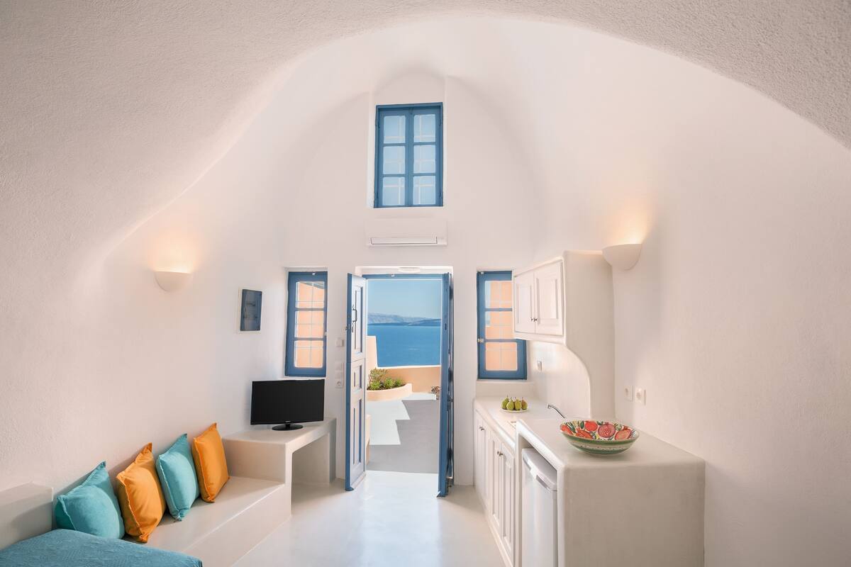 Um interior claro e branco, com uma TV sobre uma mesinha, um sofá com almofadas coloridas e uma tigela em uma bancada.