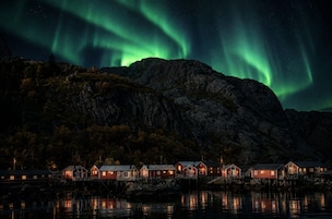 Um imóvel à beira-mar com vários edifícios vermelhos, alguns com varandas, sob um céu noturno com aurora boreal verde.