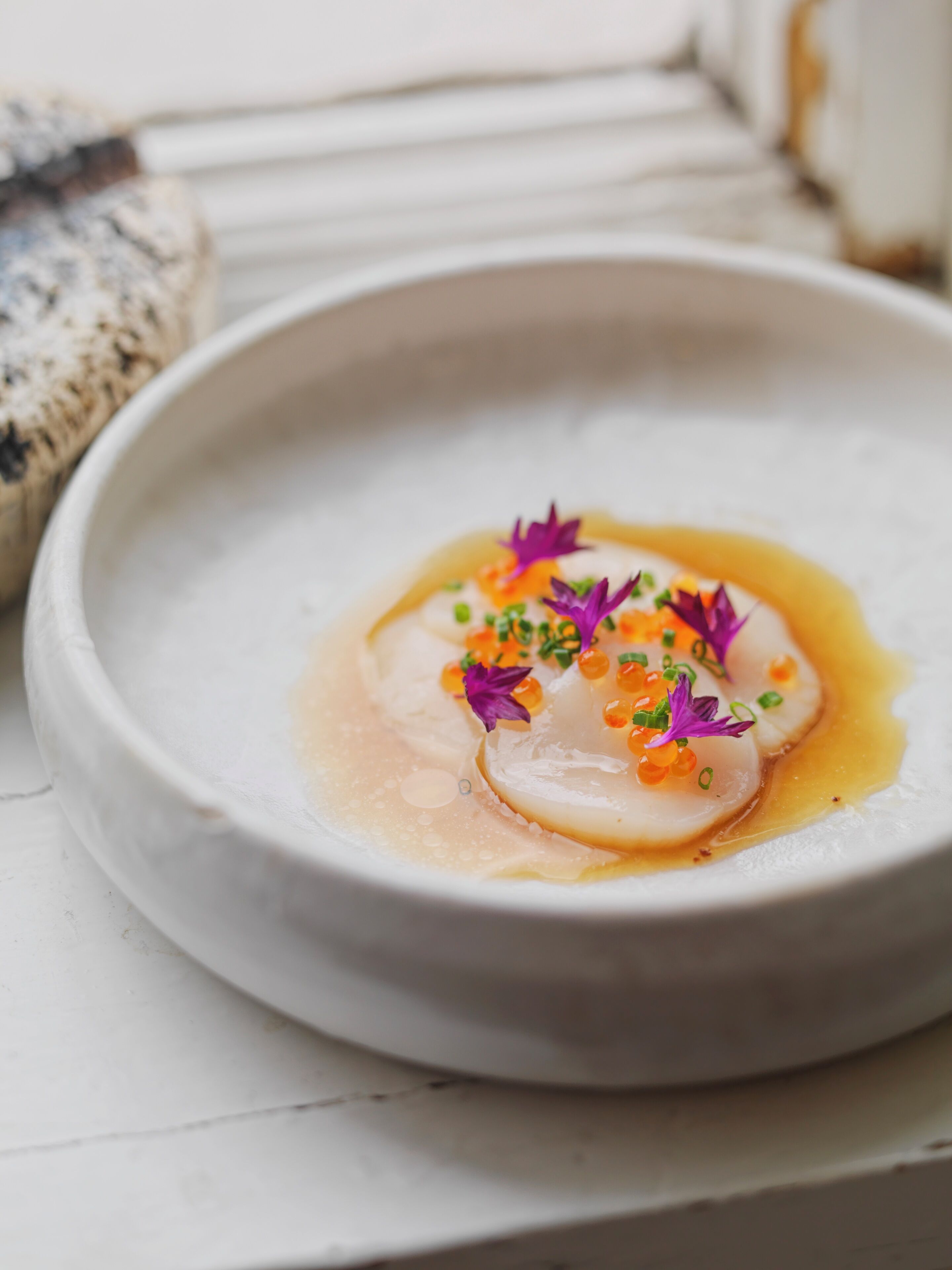 Um prato de cerâmica branco com um prato de frutos do mar, decorado com flores roxas e ervas verdes, sobre uma superfície branca.