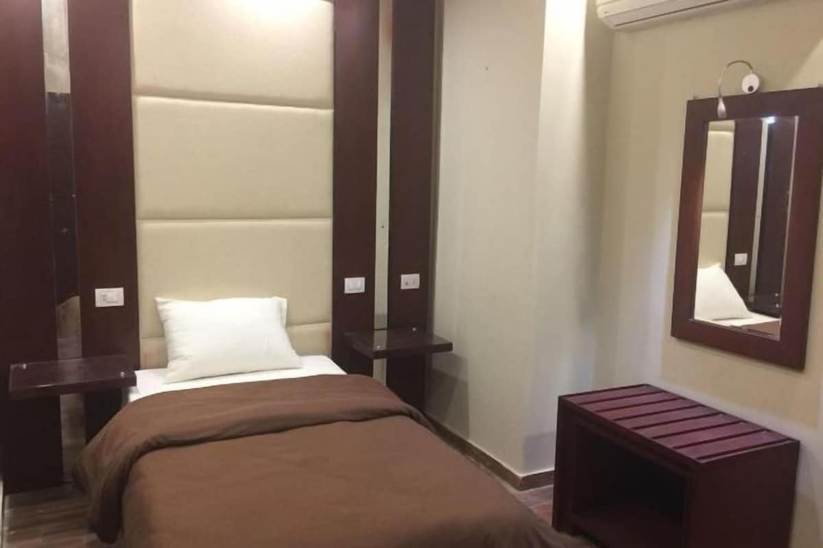 Quarto de hotel com uma cama, mesa de cabeceira, pequena mesa, espelho e ar condicionado fixado na parede.