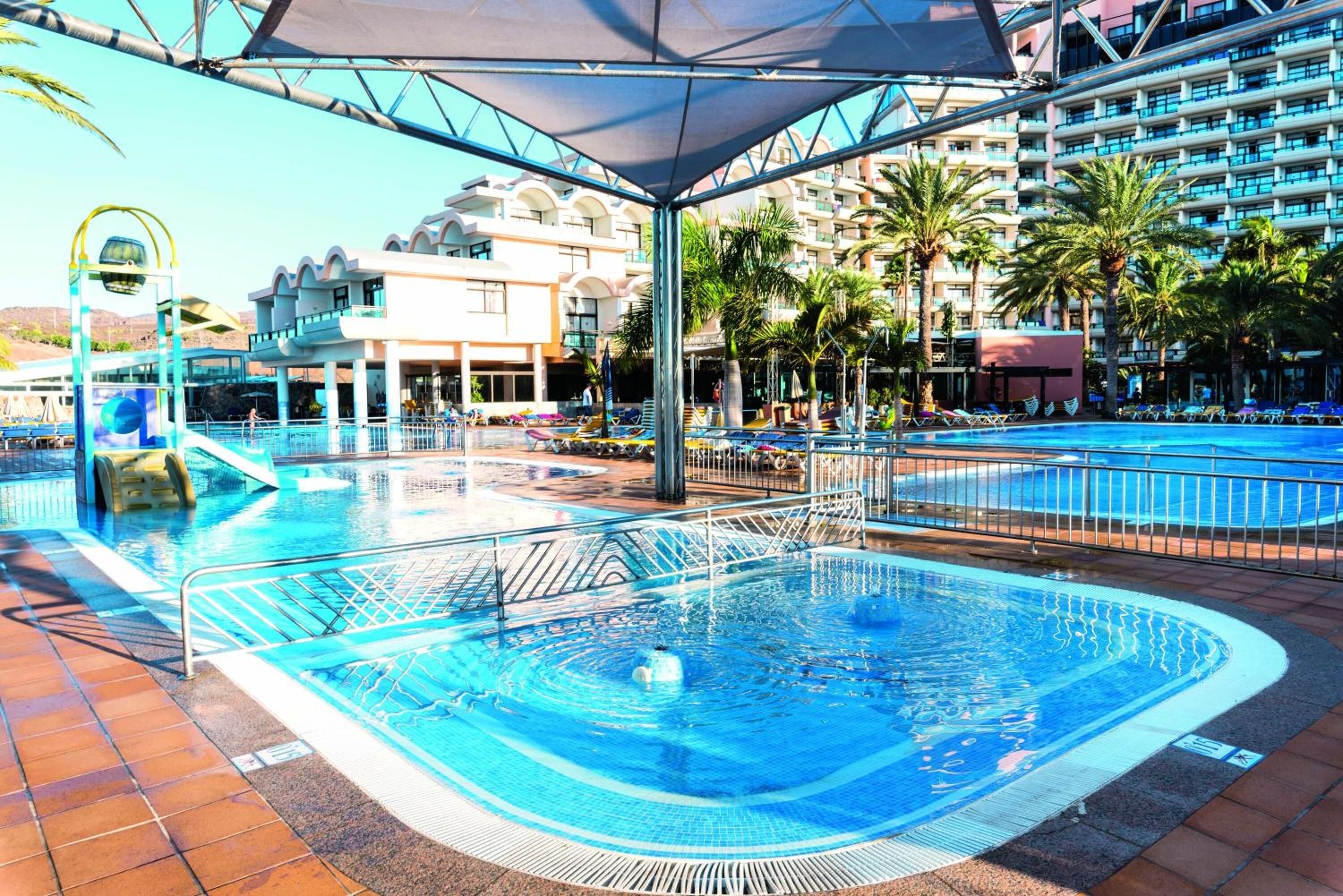Área de piscina do resort com várias piscinas, palmeiras e um espaço coberto para relaxar.