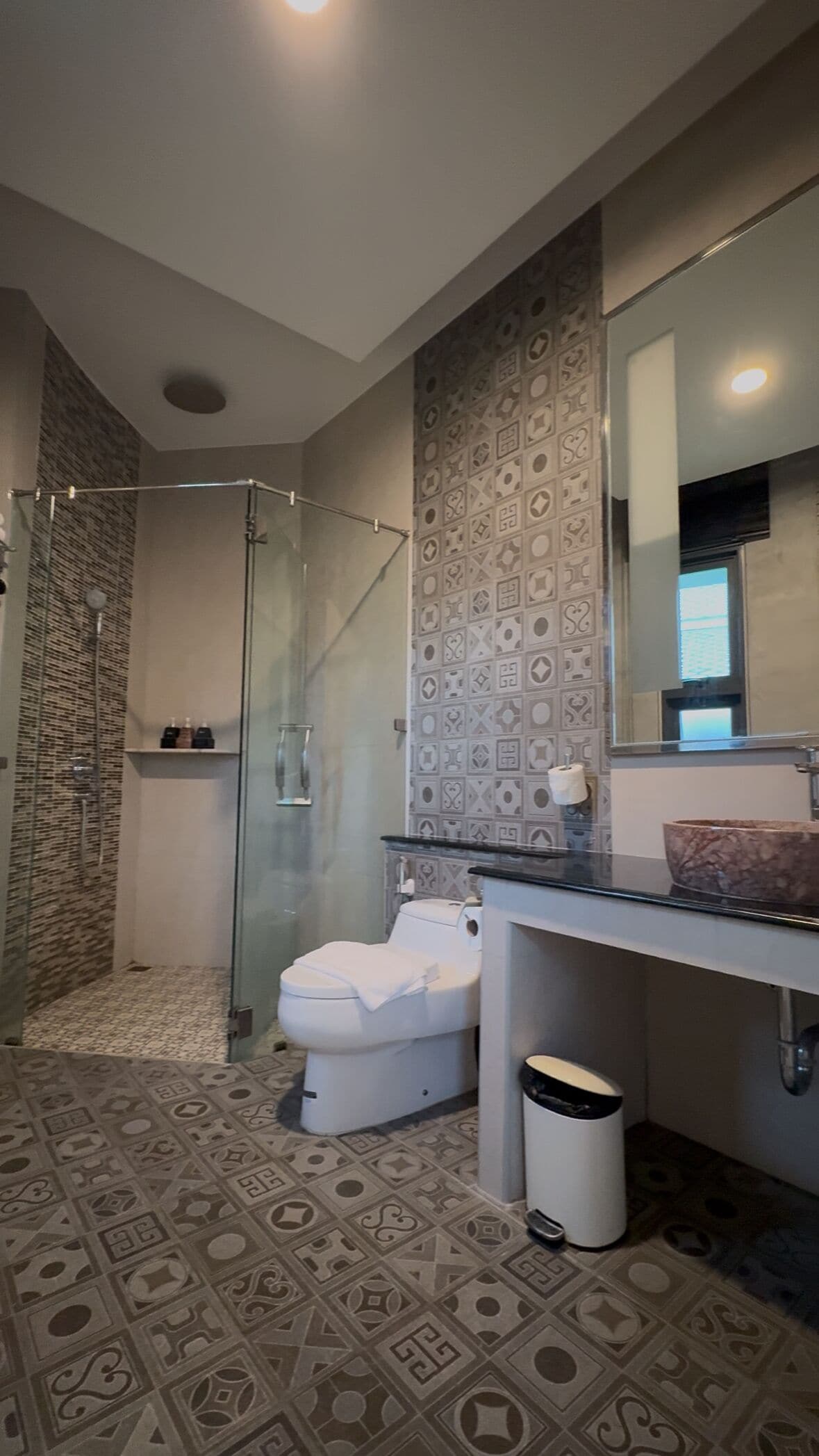 Uma casa de banho moderna com cabine de duche em vidro, sanita, lavatório com pias de mármore e pavimento em azulejos estampados.
