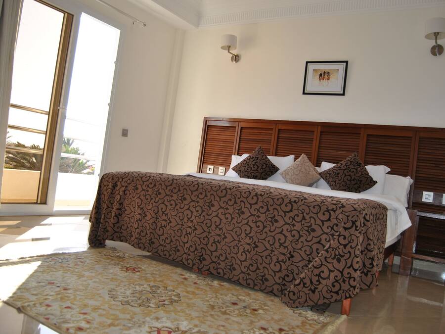 Quarto de hotel com uma cama grande, cabeceira de madeira, colcha estampada, mesa de cabeceira pequena e um quadro na parede.