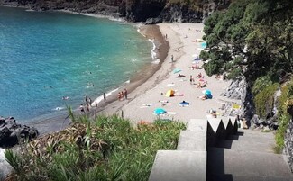 Uma praia com águas turquesa cristalinas, pessoas nadando e tomando sol, e um rochedo com vegetação.