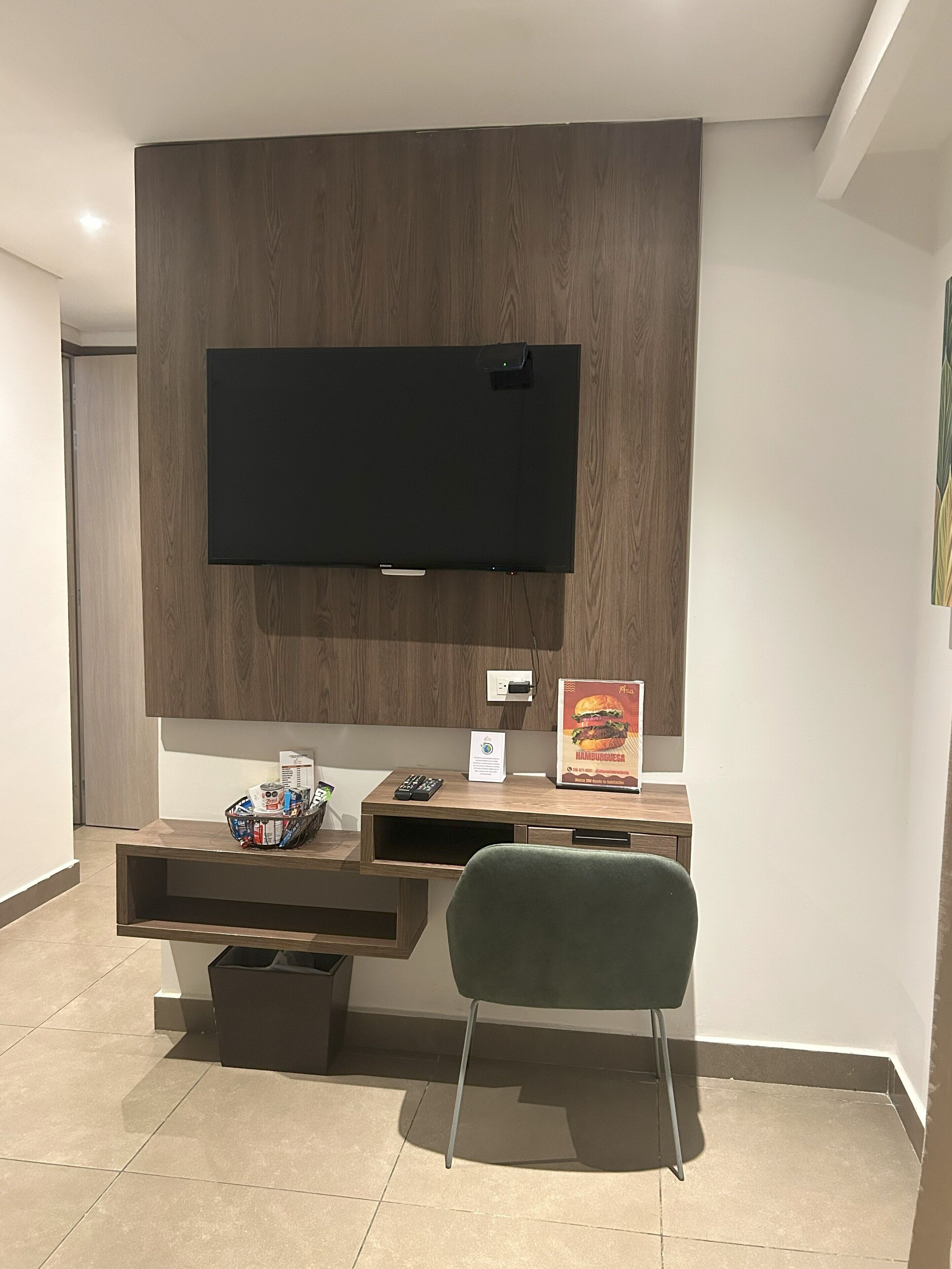 Uma sala de estar moderna com uma TV de tela plana fixada em uma parede de madeira, uma pequena escrivaninha com uma cesta de objetos, uma cadeira verde e uma lixeira.