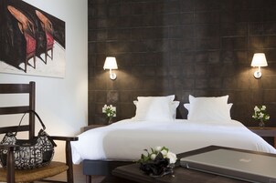 Quarto de hotel com uma cama grande, duas luminárias de cabeceira, um laptop sobre uma mesa de cabeceira, uma cadeira e uma bolsa.