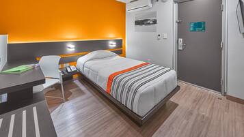 Quarto de hotel com cama, escrivaninha e cadeira. Há uma porta com uma placa e um aparelho de ar condicionado na parede.