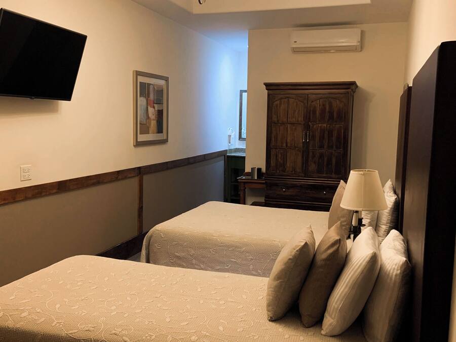 Quarto de hotel com duas camas, televisão presa na parede, lustre, armário de madeira e abajur.