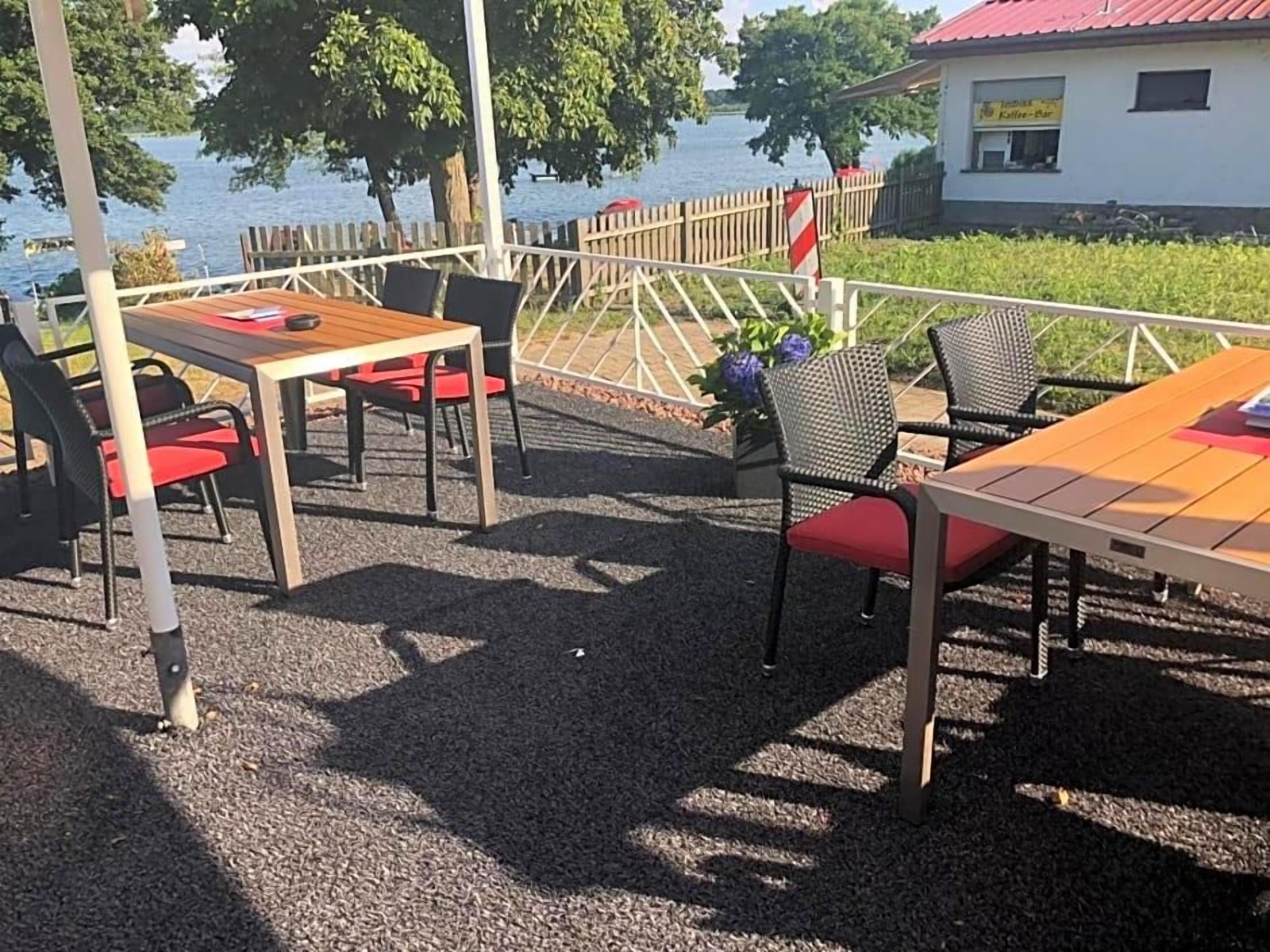 Área externa com mesas e cadeiras, vista para a água e um edifício com telhado vermelho ao fundo.