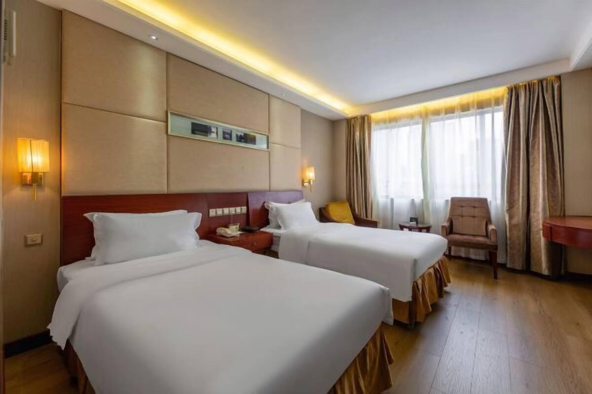 Quarto de hotel com duas camas, cabeceira de madeira, escrivaninha, uma cadeira e janela com cortinas.