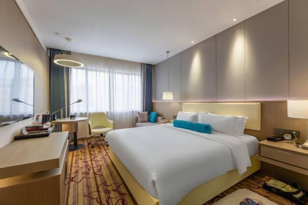 Quarto de hotel com cama grande, escrivaninha, cadeira, abajur e janela com cortinas.