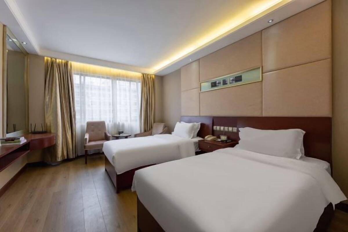Quarto de hotel com duas camas, uma escrivaninha, uma cadeira e uma janela grande com cortinas.