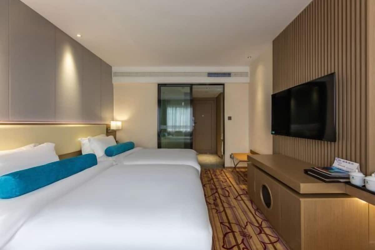 Quarto de hotel com duas camas, televisão, escrivaninha e vista para o corredor.