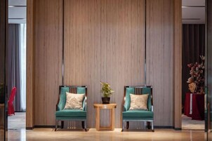 Um hall com duas poltronas estofadas em verde, uma mesinha de madeira com uma planta e um painel divisório de madeira.