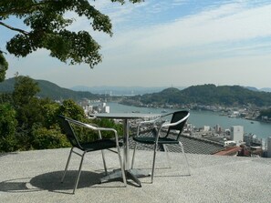 Terraço com duas cadeiras e uma mesa, com vista para uma paisagem urbana que inclui uma ponte e colinas.
